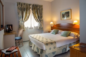 Quarto de hotel com cama, duas luminárias de cabeceira, uma cadeira, escrivaninha com luminária, quadro na parede e janela com cortinas.