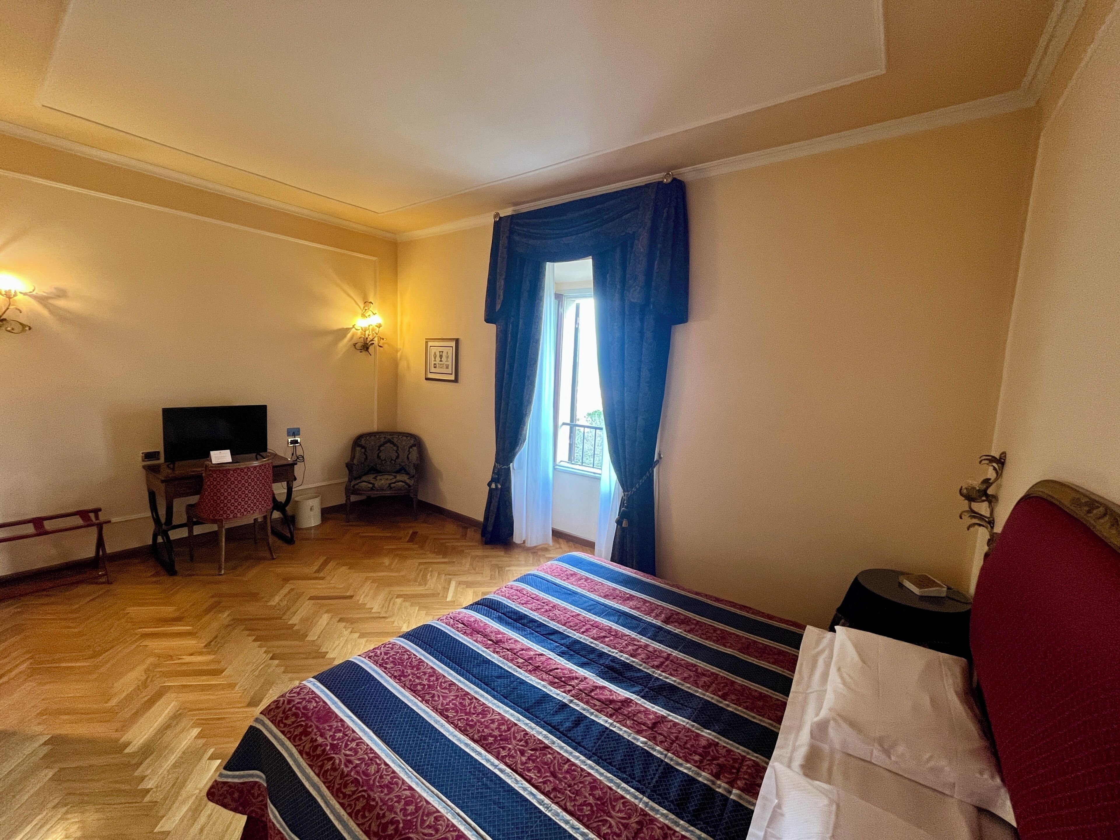 Quarto de hotel com cama, televisão, cadeira, escrivaninha e janela com cortinas.