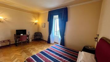 Quarto de hotel com cama, televisão, cadeira, escrivaninha e janela com cortinas.
