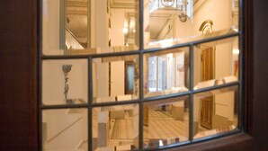Vista através de uma janela com padrão de grade, refletindo um corredor com piso de azulejos, uma porta e uma escada.