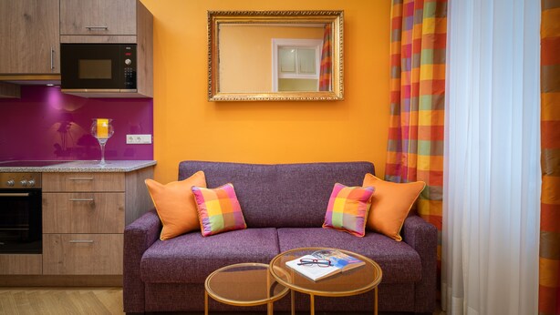 Uma sala de estar moderna com um sofá roxo, almofadas coloridas, uma mesa redonda pequena e uma área de cozinha com micro-ondas e forno.