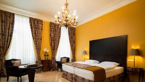 Quarto de hotel com uma cama grande, um candelabro, uma escrivaninha e uma cadeira.