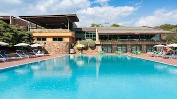Piscina com espreguiçadeiras e guarda-sóis, cercada por edifícios, uma parede de pedra e área coberta.