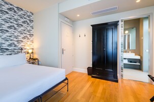 Um quarto com cama, mesa de cabeceira, guarda-roupa preto e banheiro visível por uma porta aberta.