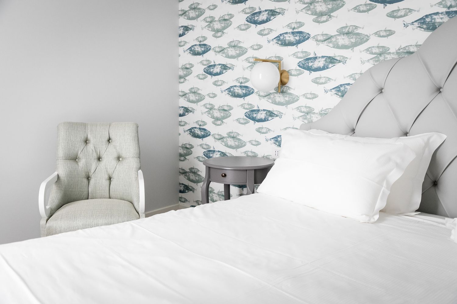 Quarto com cama, poltrona estofada, mesinha redonda e parede com padrão de peixes.
