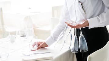 Uma pessoa segurando copos de vinho sobre uma mesa com toalha branca, copos e um caderno.