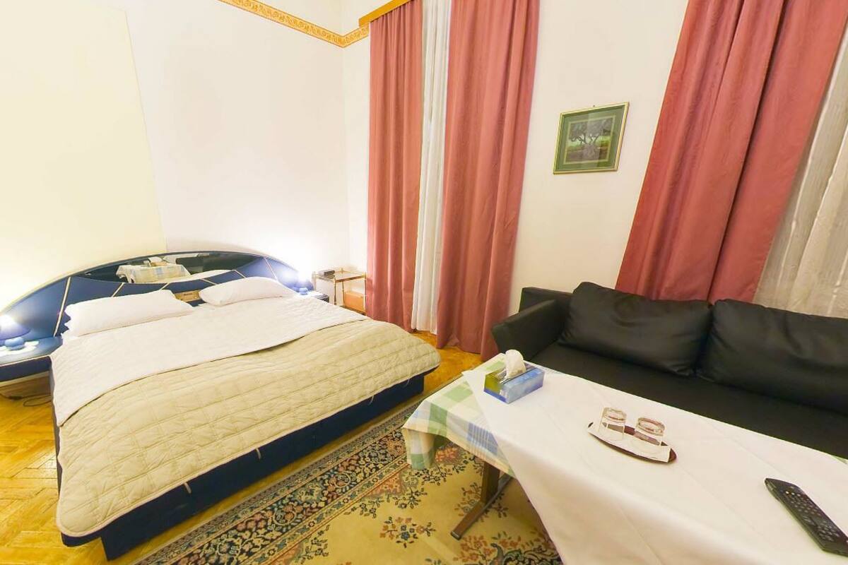 Quarto com cama, sofá preto, mesa pequena com controle remoto e tapete estampado.
