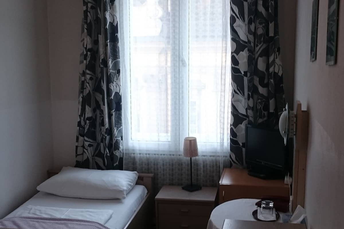 Um quarto pequeno e com pouca luz, com duas camas de solteiro, uma escrivaninha com abajur, uma televisão e uma janela com cortinas estampadas.