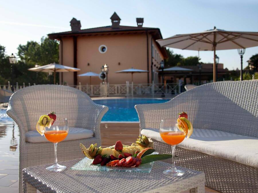 Ambiente à beira da piscina com cadeiras de vime, mesa de vidro com frutas e bebidas, e guarda-sóis.