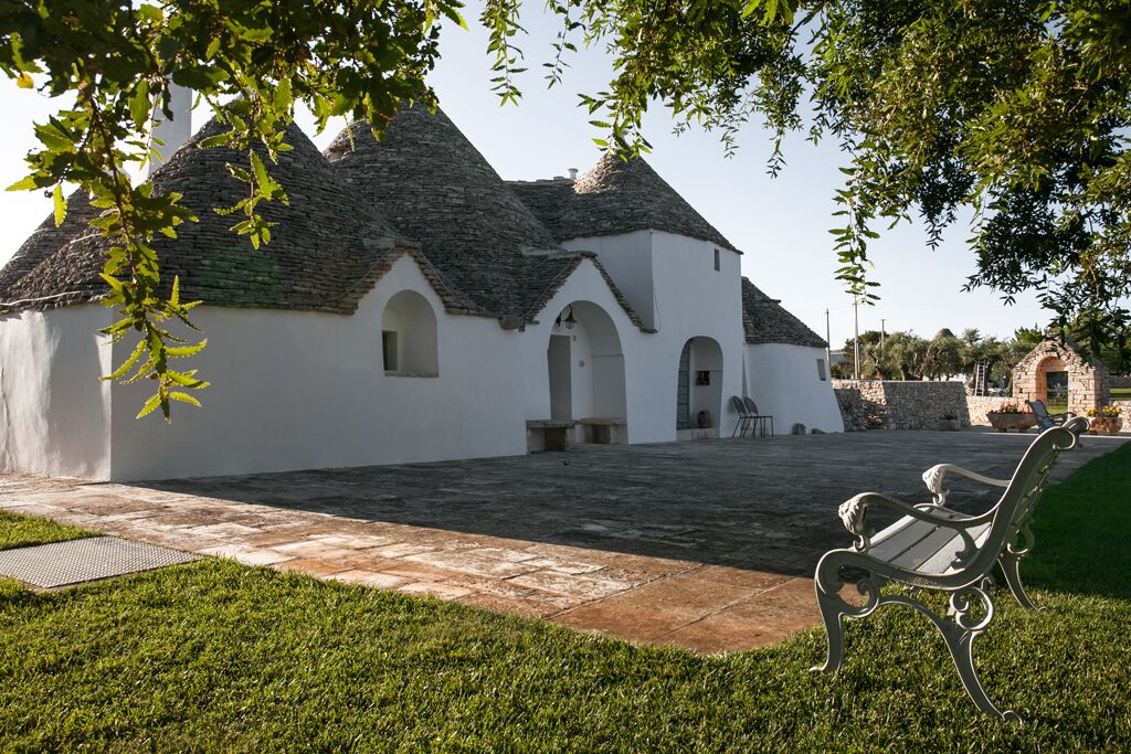 Um edifício branco com telhado de pedra e janelas em arco, pátio pavimentado e um banco.