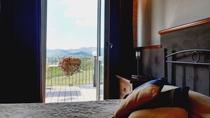 Quarto com cama, mesa de cabeceira, abajur e vista para um vinhedo através de uma porta aberta.
