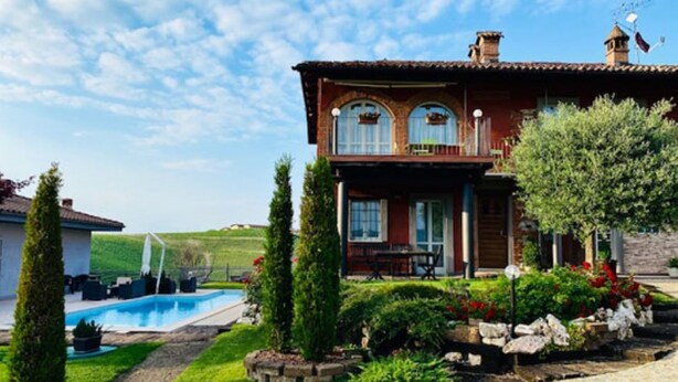 Uma casa de dois andares com piscina, um jardim com diversas plantas e flores, e um céu azul e límpido.