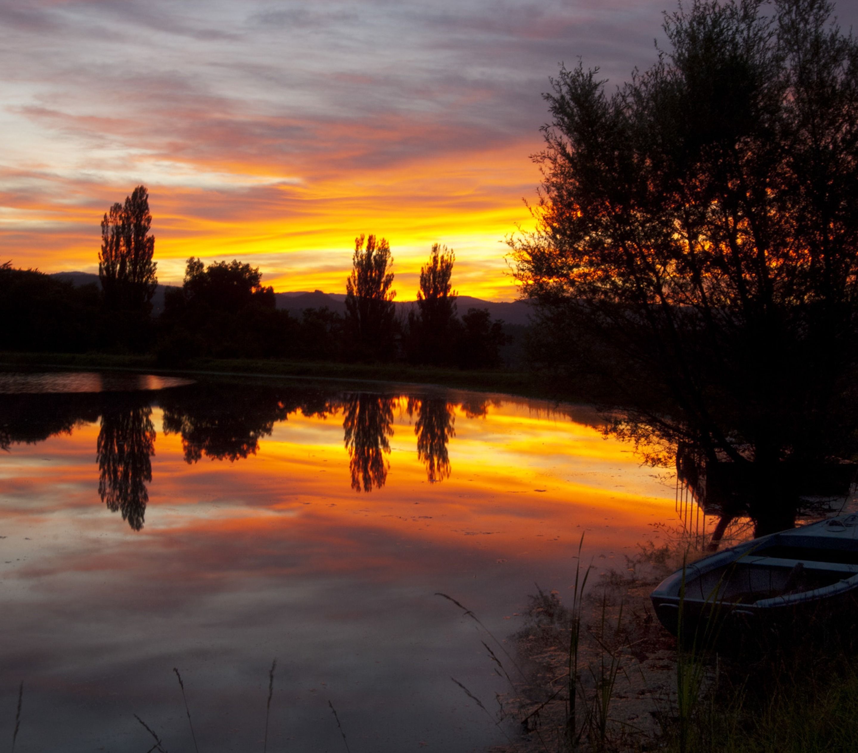 Um lago calmo refletindo um pôr do sol, com árvores e um barco à direita.