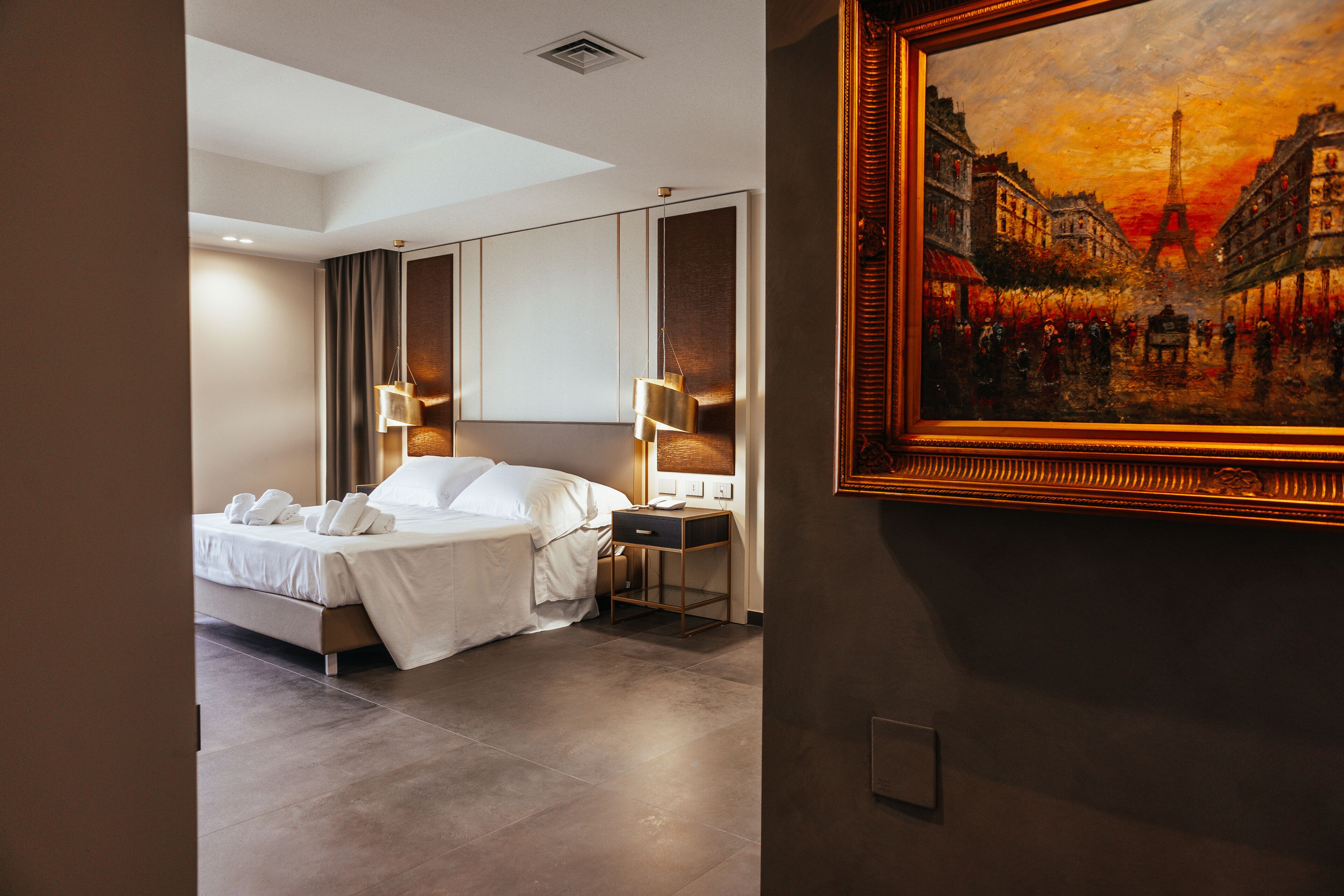 Quarto de hotel moderno com uma cama grande, mesas de cabeceira e uma pintura de uma paisagem urbana.