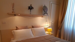Quarto de hotel bem arrumado com cama, almofadas, abajur, pequena mesa e prateleira na parede com objetos decorativos.