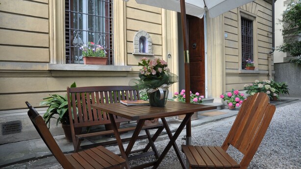 Uma área externa com assentos, incluindo uma mesa e cadeiras de madeira, rodeada por vasos com plantas e flores.