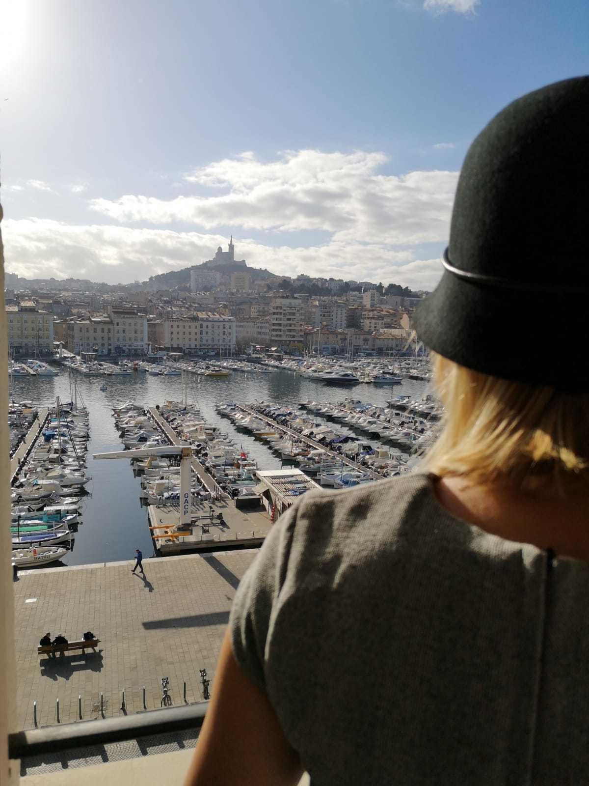 Vista de uma marina com barcos atracados, uma passarela para pedestres e edifícios ao fundo.