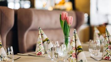 Uma mesa de jantar posta com copos de vidro transparente, um vaso com tulipas cor-de-rosa e guardanapos estampados.