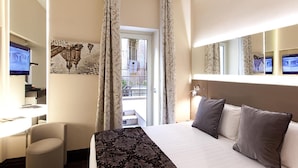 Quarto de hotel com uma cama, uma escrivaninha com computador, uma cadeira, uma janela grande com cortinas e uma televisão.