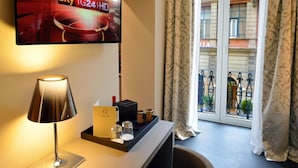 Quarto de hotel com uma televisão de ecrã plano grande exibindo 'sky TG24 HD', uma mesa de cabeceira com uma lâmpada, um tabuleiro com uma garrafa de vinho, copos e um cartão de menu.