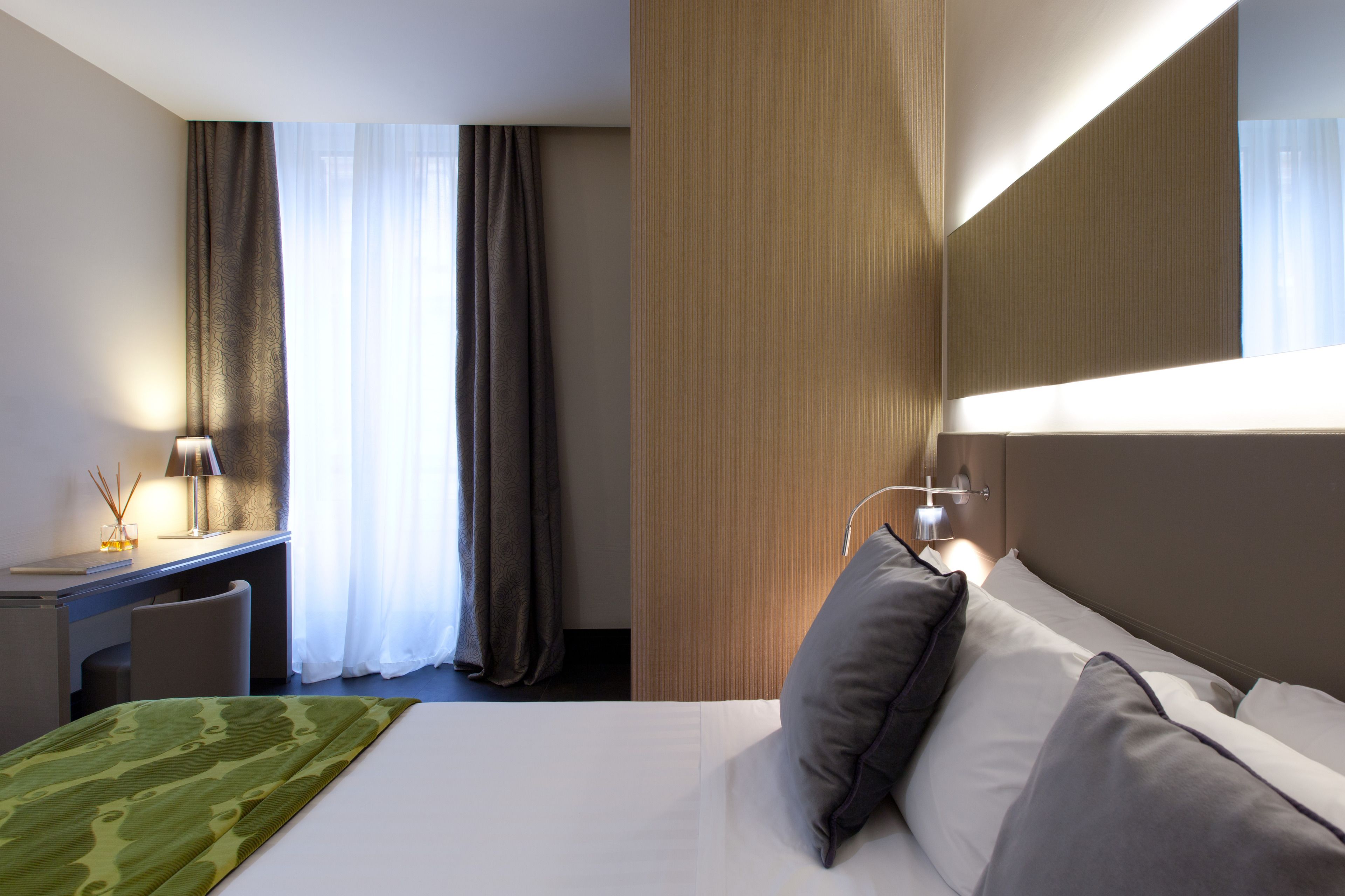 Quarto de hotel com cama, escrivaninha e cadeira. Há uma luminária na escrivaninha e uma janela com cortinas.
