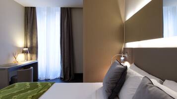 Quarto de hotel com cama, escrivaninha e cadeira. Há uma luminária na escrivaninha e uma janela com cortinas.