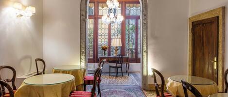 Uma sala de jantar com mesas e cadeiras redondas, um candelabro e vista através de uma janela com vitrais.