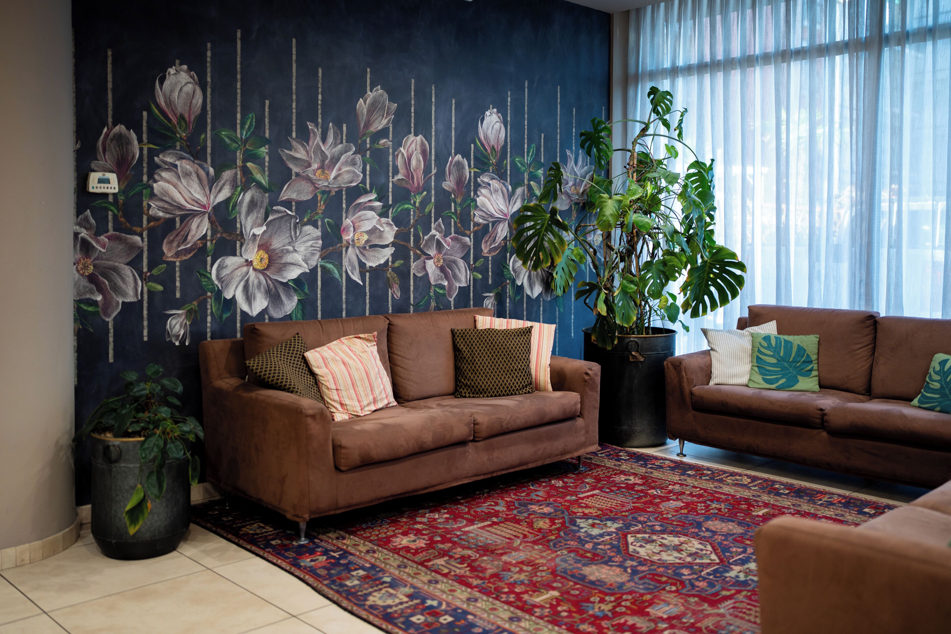 Sala de estar com dois sofás, uma grande planta em vaso, tapete estampado e parede com um mural floral.