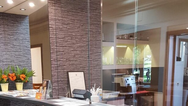 Balcão de recepção com design moderno, destacando-se por um balcão de madeira e um painel de vidro. Há plantas em vasos e um certificado emoldurado no balcão.