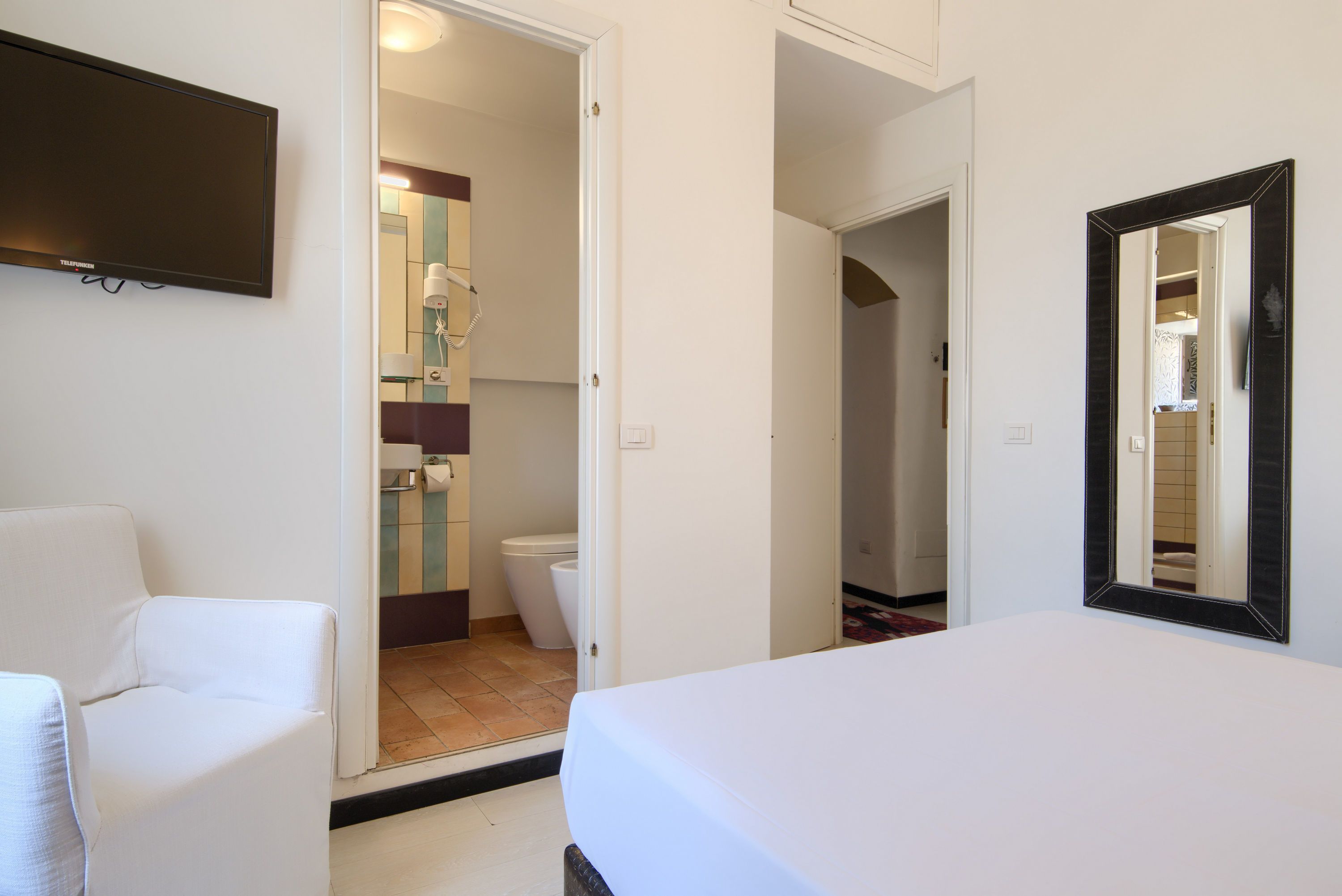 Quarto de hotel com cama, televisão na parede, poltrona branca e banheiro visível através de uma porta aberta.
