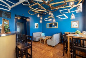 Interior de restaurante com parede azul, teto de madeira e luminárias pendentes. Há mesas e cadeiras de madeira, um balcão com menu em lousa e um sofá.