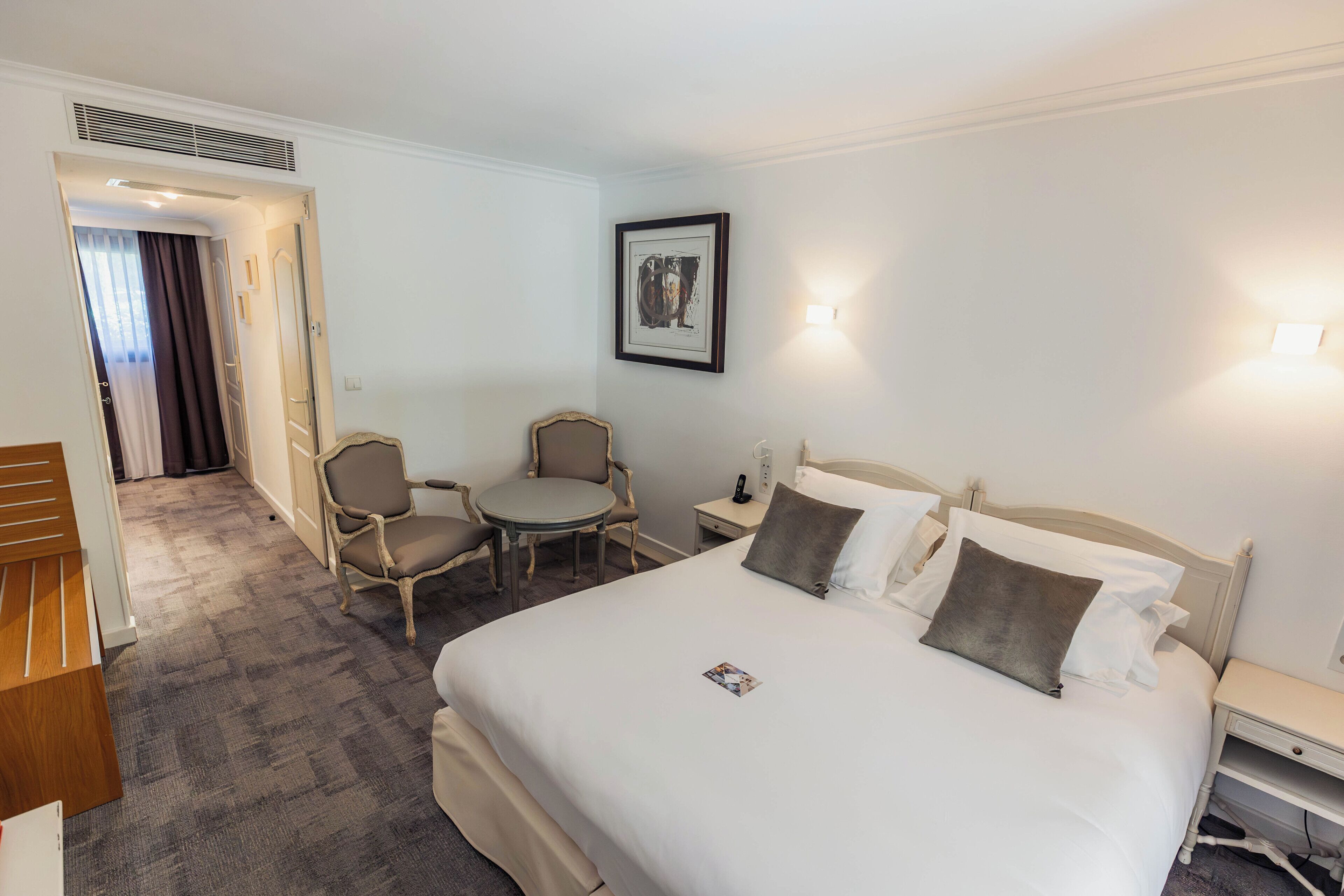 Quarto de hotel com uma cama grande, uma mesa redonda pequena, duas poltronas, uma mesa de cabeceira e um quadro emoldurado na parede.