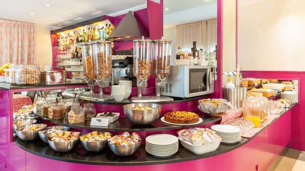 Uma área de buffet com diversos alimentos e bebidas, incluindo tigelas de cereais, doces e uma saladeira de frutas.