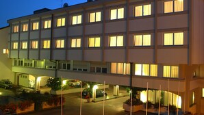 Um hotel com vários andares, janelas iluminadas, área de estacionamento e plantas ao ar livre.