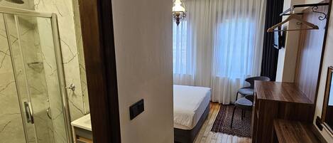 Quarto de hotel com piso de madeira, uma cama, uma mesa de trabalho, chuveiro com portas de vidro e vista para o exterior através de uma janela com cortinas.