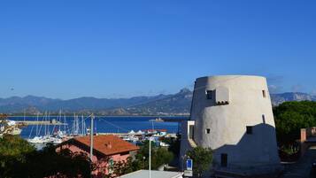 Uma vila costeira com uma torre imponente, marina com barcos e montanhas ao fundo.