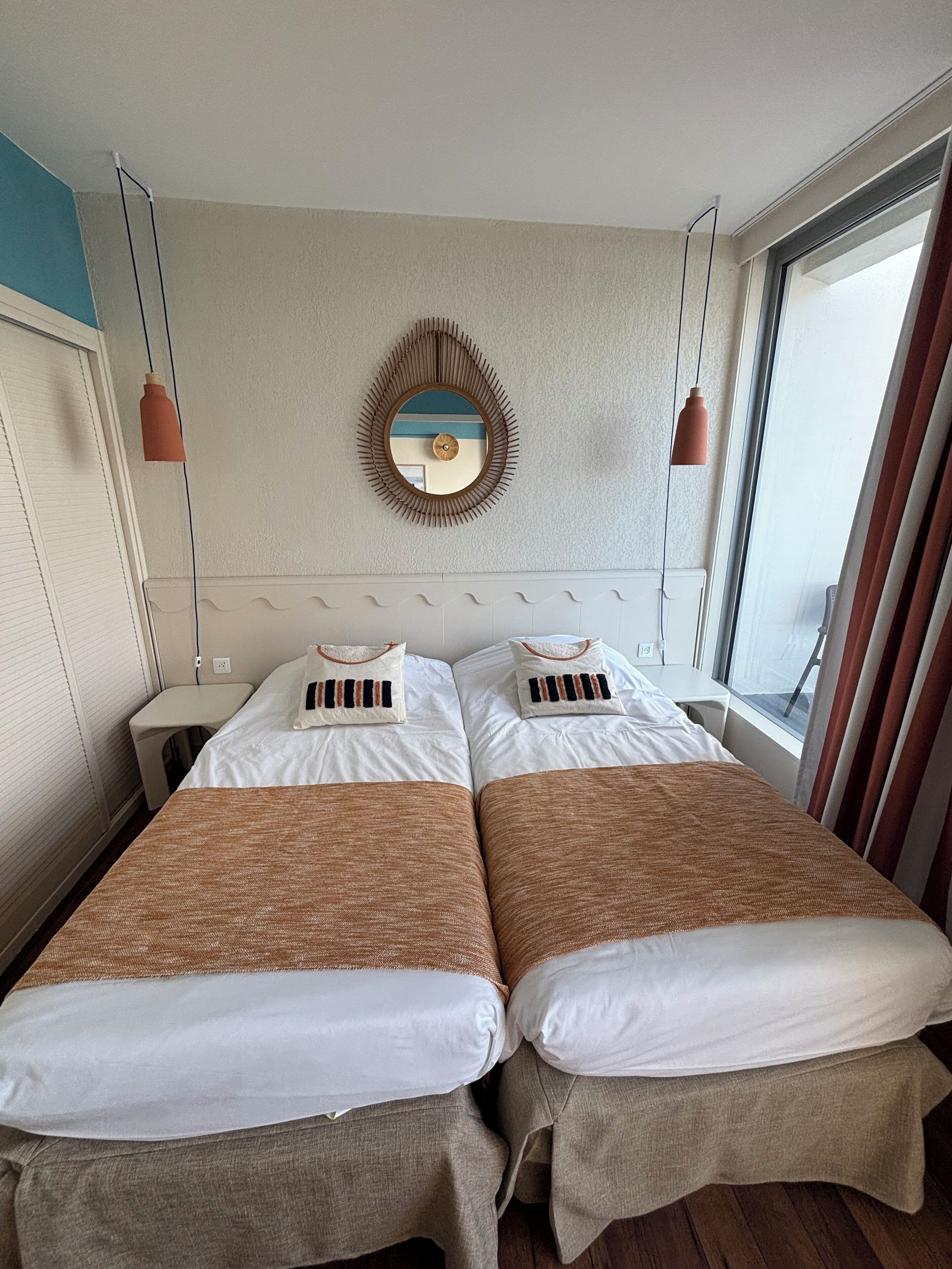 Quarto de hotel com duas camas, um espelho redondo, luminárias pendentes e uma janela com cortinas.