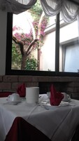 Mesa de jantar posta para dois, com toalhas brancas e guardanapos vermelhos, uma chávena branca e uma tigela branca. Uma janela mostra uma árvore com flores cor-de-rosa e um edifício ao fundo.
