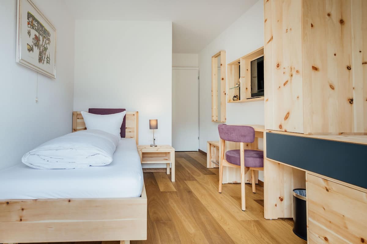 Quarto minimalista com cama de madeira, escrivaninha, cadeira e televisão fixada num armário de madeira.