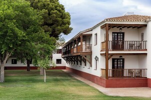 Um edifício de dois andares com uma varanda, uma árvore em primeiro plano e céu claro.