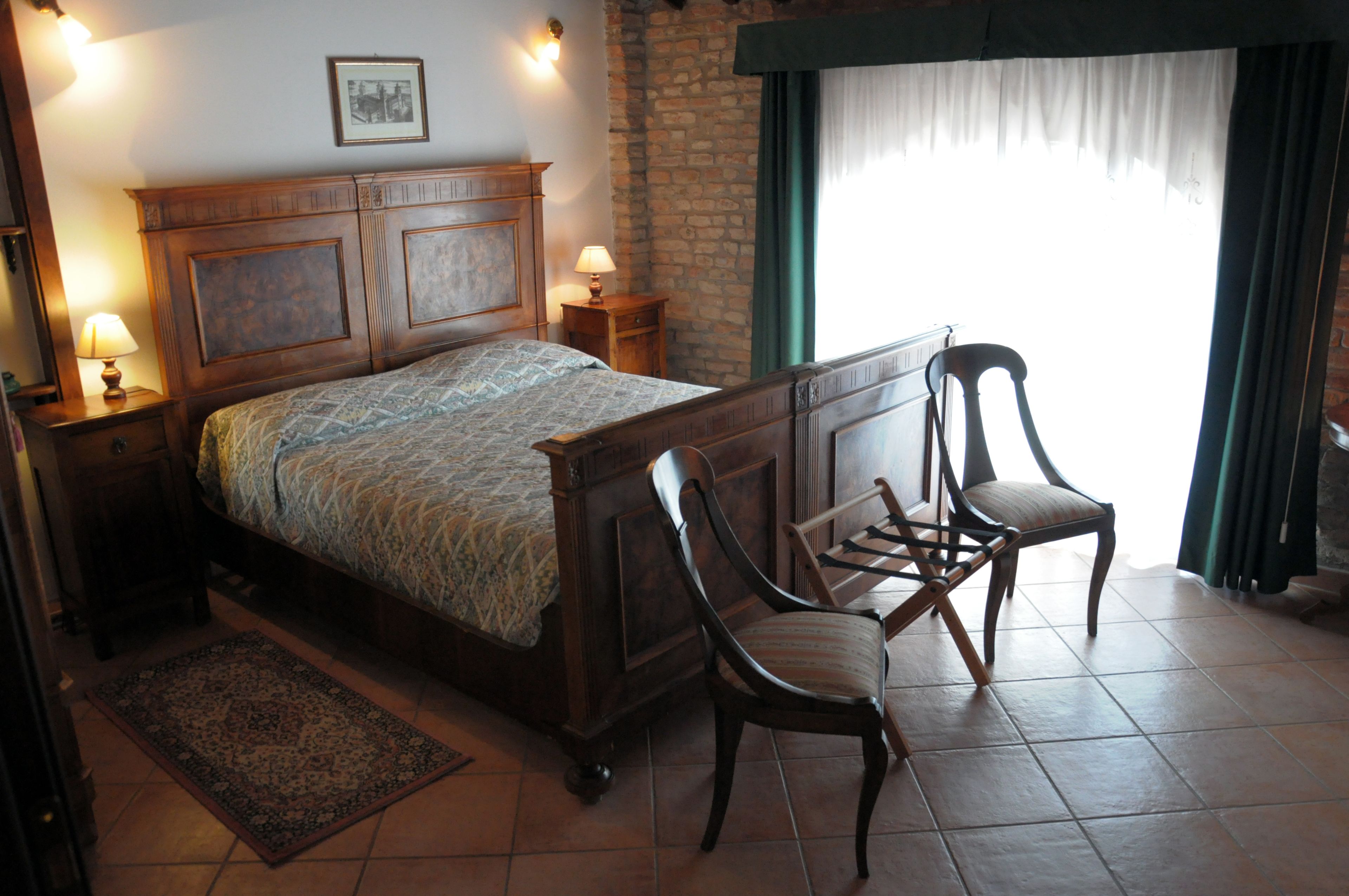 Quarto com uma cama grande, duas cadeiras, uma mesa de cabeceira com abajur, um quadro na parede e uma janela com cortinas.
