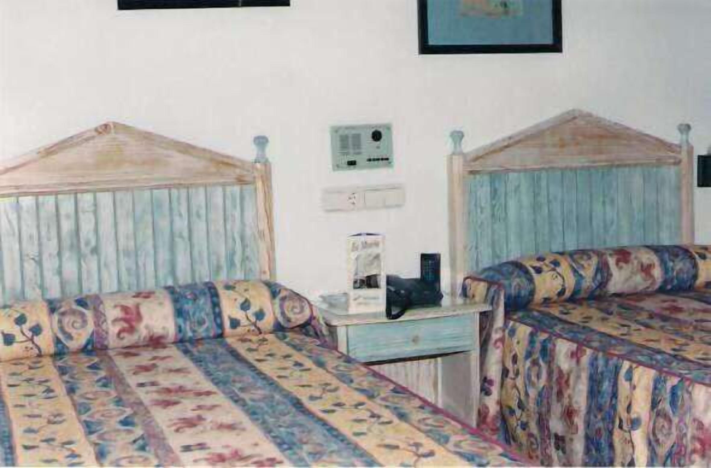 Quarto com duas camas com roupa de cama estampada, uma mesinha de cabeceira com abajur e um termostato fixado na parede.