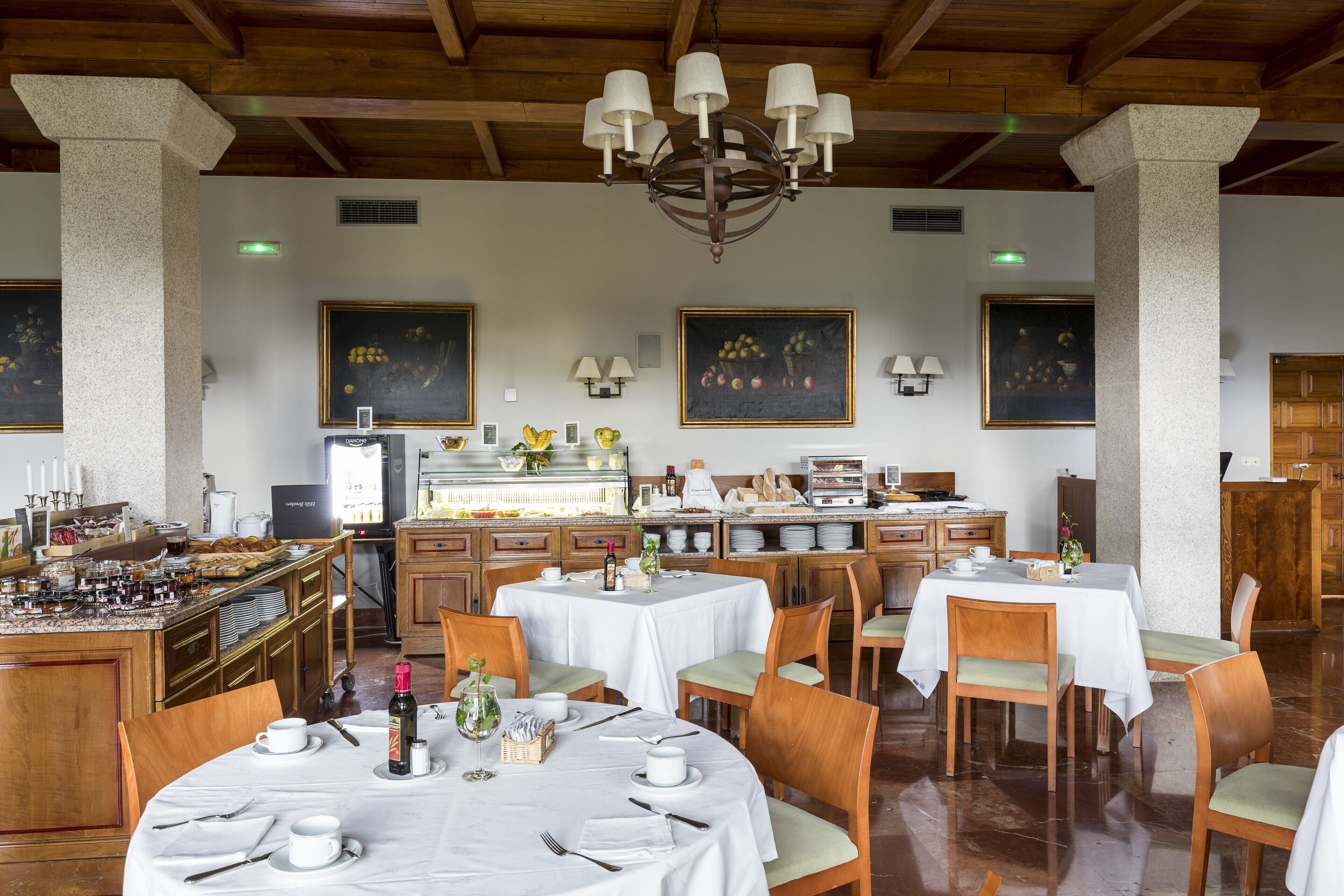 Uma área de refeições com várias mesas e cadeiras, uma ilha central com comida e bebidas, e quadros nas paredes.