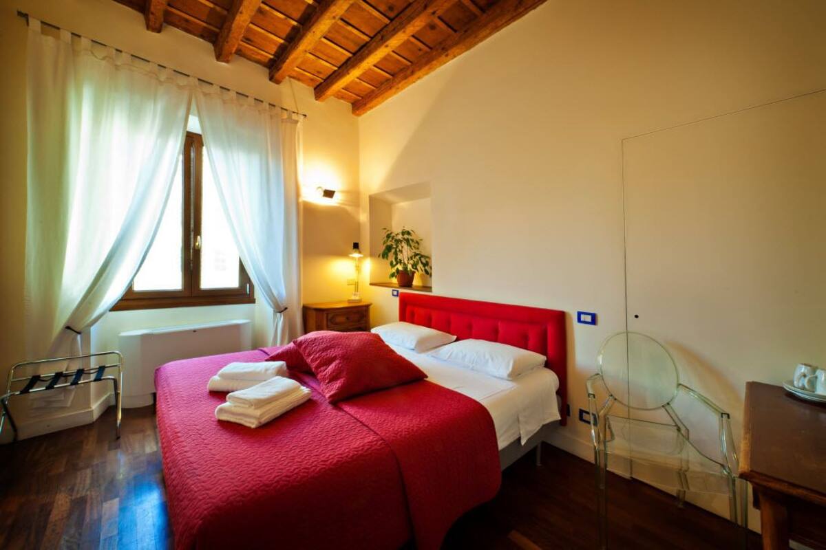 Quarto de hotel com teto de madeira, cama com roupa de cama vermelha, mesa de cabeceira com abajur, uma cadeira e janela com cortinas.