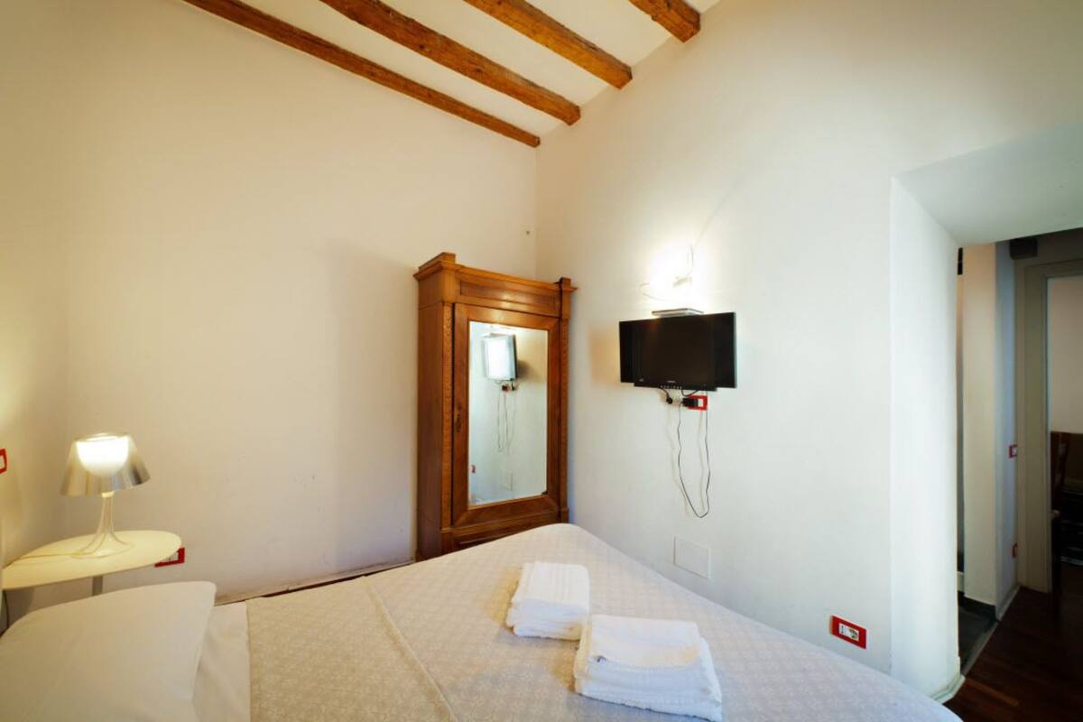 Quarto de hotel com uma cama, um guarda-roupa de madeira, uma TV fixada na parede e um abajur.