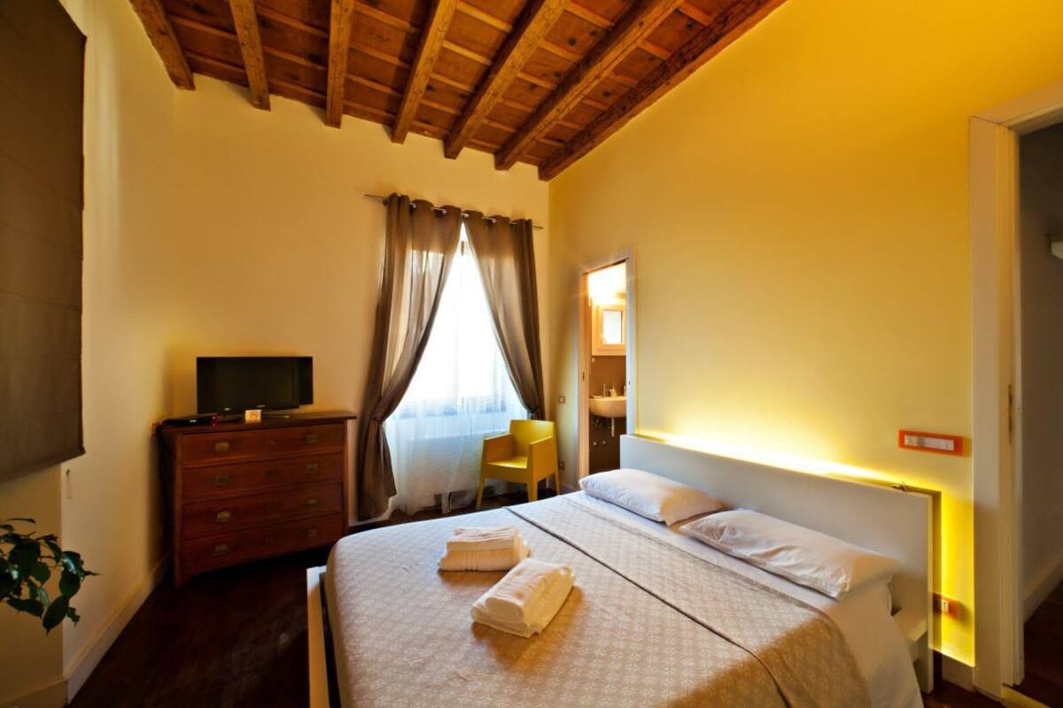Quarto de hotel com teto de madeira, cama com lençóis brancos, televisão em uma cômoda e janela com cortinas.