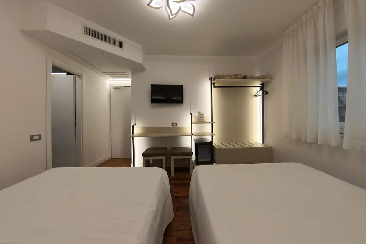 Quarto de hotel com duas camas, uma escrivaninha com cadeira, televisão e janela com cortinas.