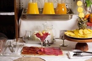 Um buffet de café da manhã com fatias de carne, uma pinha como peça central e jarras de suco de laranja.