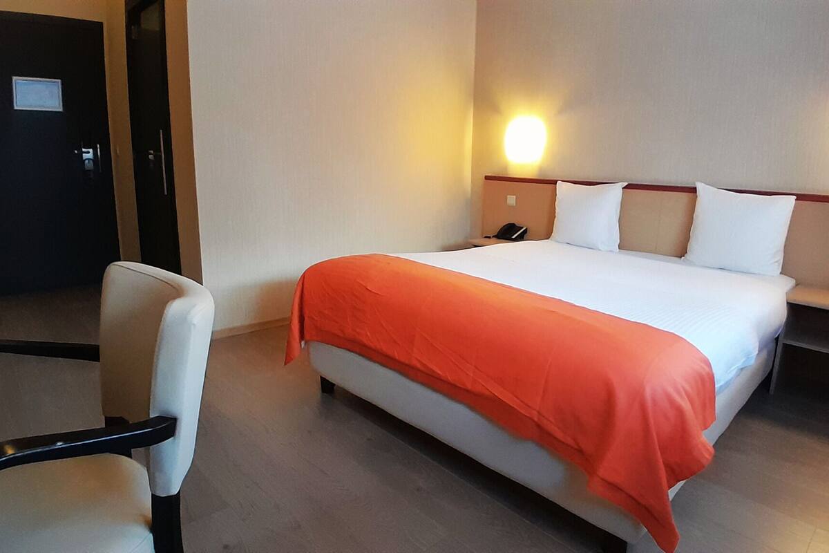 Quarto de hotel com cama, cadeira, mesa de cabeceira e telefone fixado na parede.