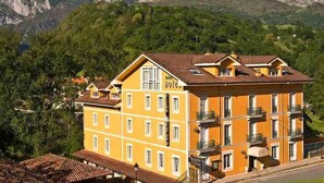 Um edifício amarelo com várias varandas, uma placa indicando que é um hotel, e montanhas ao fundo.